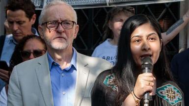 Jeremy Corbyn and Zarah Sultana stand among a group of people at a protest. Corbyn wears a light-colored blazer over a blue shirt. Sultana, dressed in a black outfit with colorful embroidery, holds a microphone. 