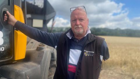 A bald man with a beard hangs from the side of a yellow tractor on a farm. His other arm is missing with the shirt sleeve flapping loose