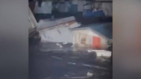 Two small houses float in water that is littered with debris. One has an orange door and a blue roof.
