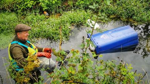 Man in green jacket, flat cap, hi-vis vest and red gloves waist-deep in water surrounded by leaves and branches. He is holding a plastic bag of rubbish and moving towards a blue wheelie bin on its side in the water. 
