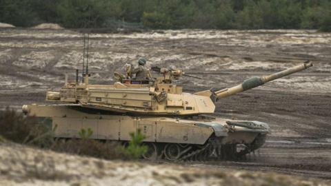 A sand-coloured M1A2 Abrams tank manned by a US soldier from the 2nd Battalion during an exercise in Poland in 2023. The tank is standing in a slushy landscape, with a deep green tree-line visible in the distance. 