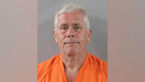 A man in his sixties with white hair looks at the camera during his police mug shot.
