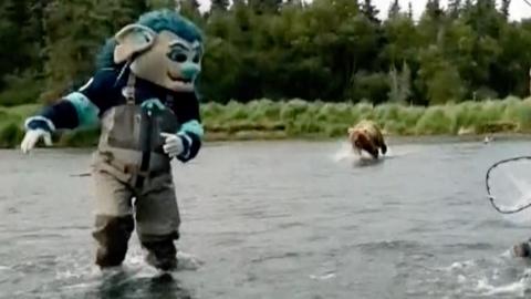 Someone dressed as Buoy the NHL mascot and wearing high waders, is seen running in knee deep water while being chased by a bear. Another man wearing fishing gear is also fleeing the running animal. In the background is a forest. 