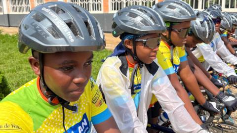 Several young female cyclists, wearing grey cycle helmets, bright jerseys and a smattering of sunglasses and see-through raincoats, line up on their bikes. The photograph is taken looking down the line, with the nearest cyclist's face seen close up in detail