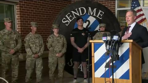 A screenshot of a press conference held by the US army on Thursday. In it, US Army Secretary Dan Driscoll is seen speaking at a wooden podium, with several microphones in front of him. Six soldiers are seen standing on his left, with five of them in full Army uniform and one wearing black shorts and a T-shirt. A US flag on a pole is also seen behind him.