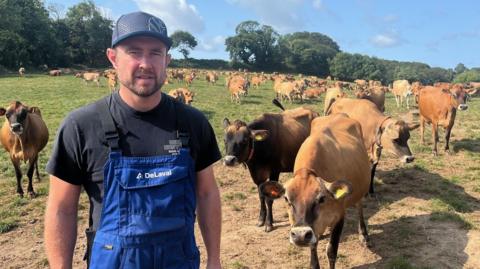 Tom Perchard is standing in a field surrounded by his Jersey cows on a sunny day. The grassy field behind him is covered in cows and the ones in the foreground close to him are looking at the camera. They are wearing collars and have ear tags in them. Mr Perchard is wearing a pair of dungarees over a T-shirt and a cap. He has short dark hair and a goatee.
