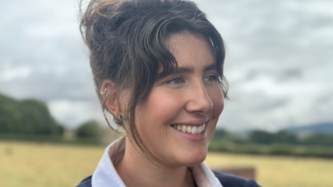 A woman with brown hair pulled into a bun smiles to the side of the camera. She wears silver earrings. The photo is a close-up so you can see her head and shoulders. The background is blurred but you can see the yellow crops on a farm and trees behind her.