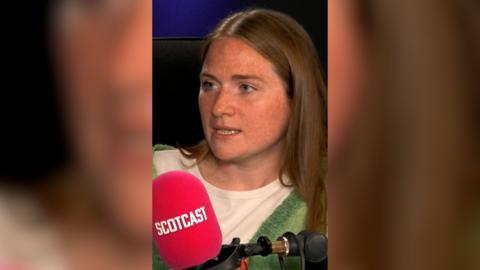 A woman with brown hair and blue eyes, wearing a white top and a green cardigan, is talking into a red 'Scotcast' mic.