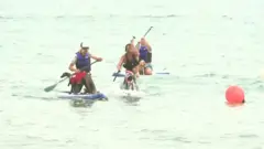 u vodi na tri daske za surf ili SUP vide se tri takmičara koji veslaju zajedno sa njihovim psima koji stoje na daskama