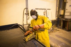 Un homme dans une combinaison jaune, un cahce-nez et des gants en train de taper sur un lingot d'or avec du marteau.