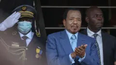Le président camerounais Paul Biya applaudit lors du défilé du 20 mai marquant la 51e célébration de la fête de l'unité à Yaoundé, le 20 mai 2023. Il porte un costume bleu et une cravate. Un soldat salue derrière lui.