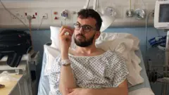 Chris Brookes-Smith w szpitalnej koszuli w szpitalnym łóżku podczas badania klinicznego