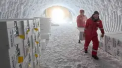 two people in sub zero protective red jump suits carry a large white chest between them through an icy tunnel towards the camera. Lots more similar boxes are stacked on each side of the walls of the cave.