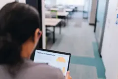 Mulher, de óculos e cabelos pretos, aparece de costas trabalhando em um tablet