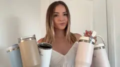 Una mujer joven con pelo largo claro, de camisa blanca, sostiene cinco recipientes Stanley 