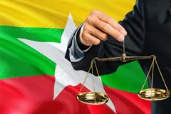 တရားစီရင်မှုနဲ့ ရှေ့နေကို ကိုယ်စားပြုတယ်လို့ မှတ်ယူနိုင်တဲ့ ကြေးသမာဓိချိန်ခွင်ကို တိုက်ပုံ အနက်ရောင်ဝတ်ထားတဲ့အမျိုးသားတစ်ဦးက ကိုင်ဆောင်ထားတဲ့ပုံကို အဖြူ အဝါ အစိမ်း အနီရောင် အလံနောက်ခံနဲ့ မြင်တွေ့ရတာဖြစ်ပါတယ် 
