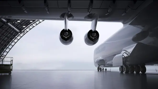 Arte conceptual enfoca el enorme avión WindRunner desde una de sus alas, mostrando dos turbinas y el fuselaje. El avión está dentro de un hangar abierto que deja ver un cielo gris
