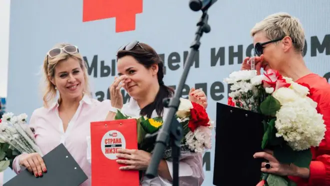 Слева направо: Вероника Цепкало в белой блузке, Светлана Тихановская с папкой "Страна для жизни" и букетом цветов, Мария Колесникова с цветами и в темных очках - на сцене предвыборного митинга в 2020 году