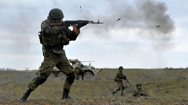 平地で銃を構える兵士の後ろに、戦車が見える