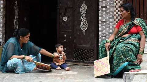 Two women holding hand fans, and a baby, sit on the street