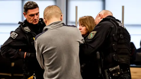 A man is seen from behind standing in front of three prison officers. 