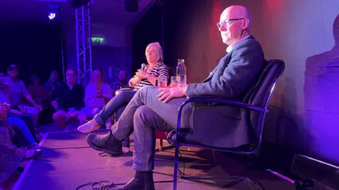 John Swinney and Susan Morrison, both dressed casually, on stage. the lighting is very purple.