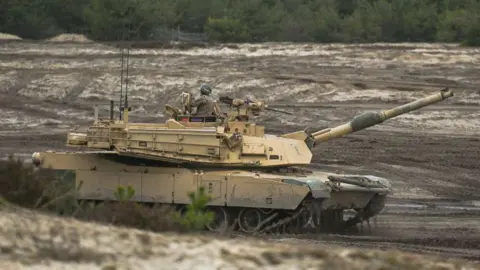 A sand-coloured M1A2 Abrams tank manned by a US soldier from the 2nd Battalion during an exercise in Poland in 2023. The tank is standing in a slushy landscape, with a deep green tree-line visible in the distance. 