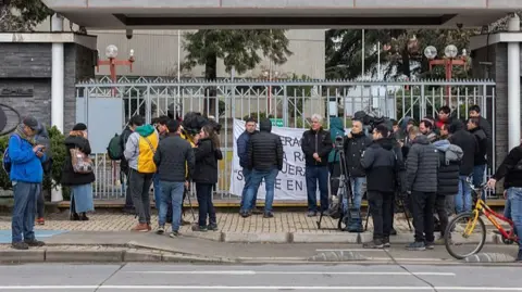 Relatives of the trapped and injured miners wait for information at Codelco HQ. They wear jeans and puffa jackets, one man has a camera.