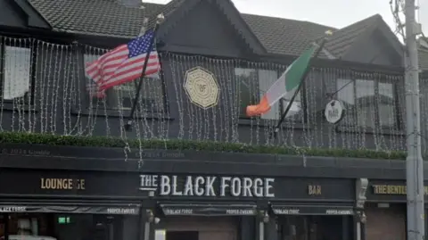 The exterior of The Black Forge pub. The building is painted black with the Black Forge written in white across the entrance. There is an Irish and a US flag flying above the entrance. 