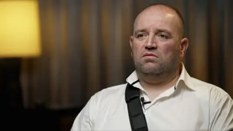 A middle aged man with a bald head and short beard wearing a white shirt sits in front of a blurred curtain